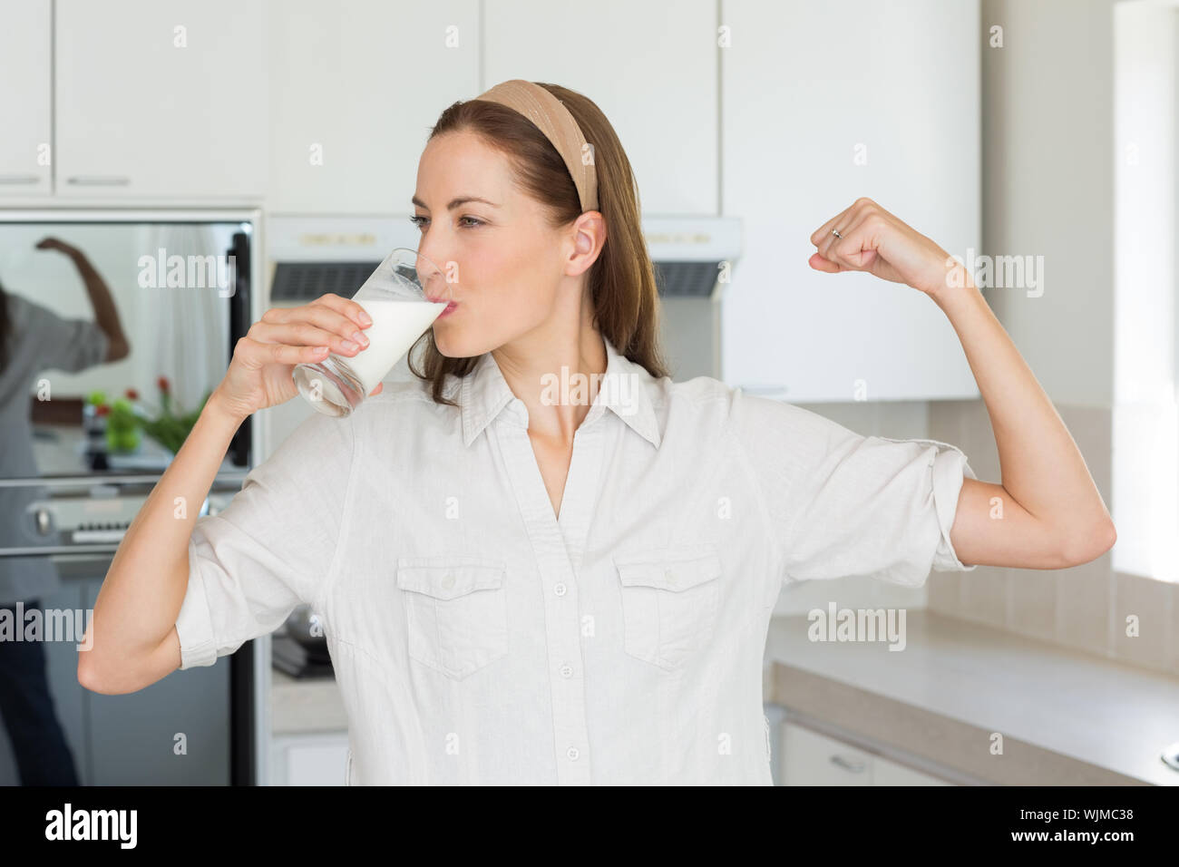 Young woman flexing muscles while drinking milk in the kitchen at home ...