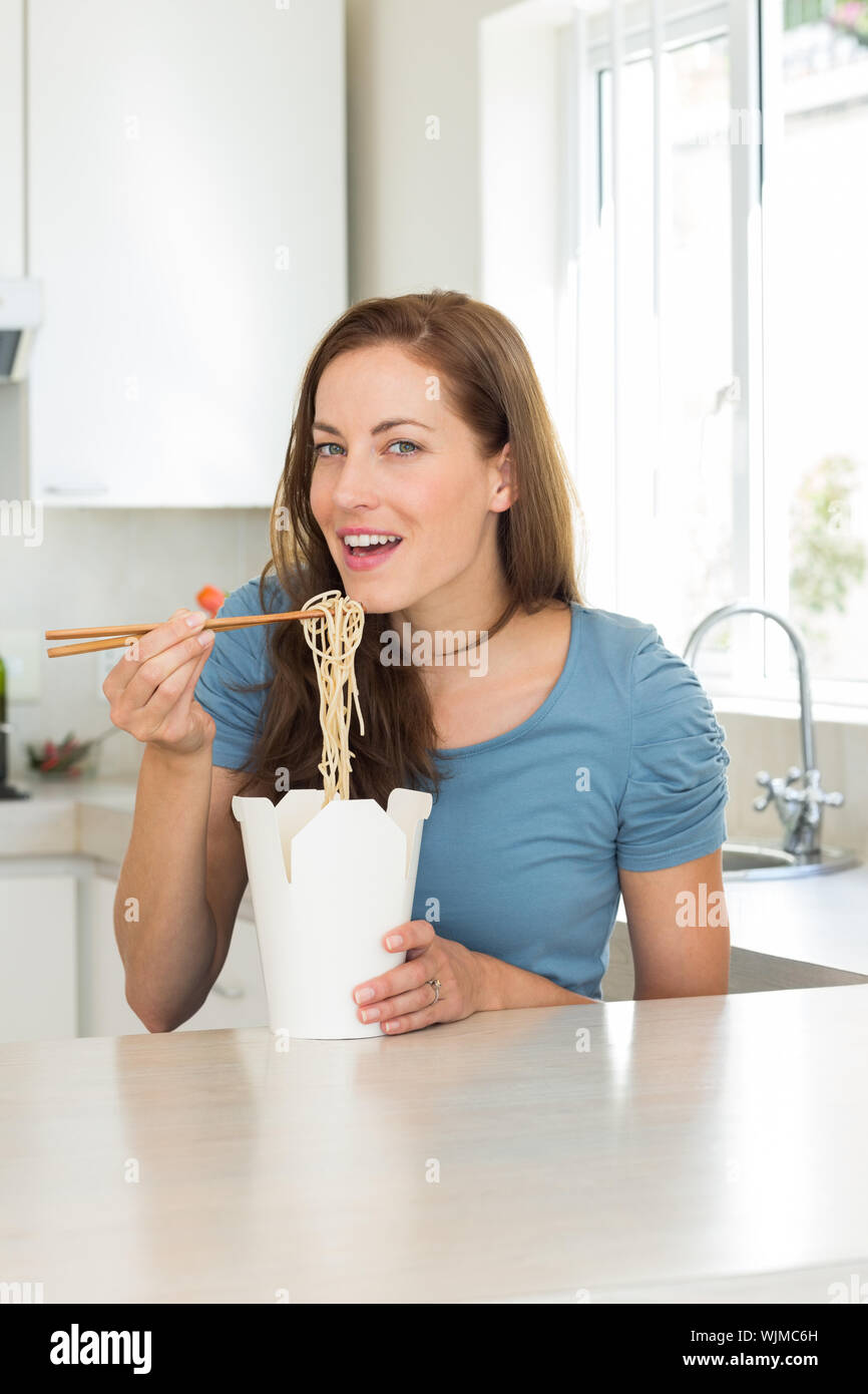 Portrait of a smiling young woman eating noodles in the kitchen at home ...
