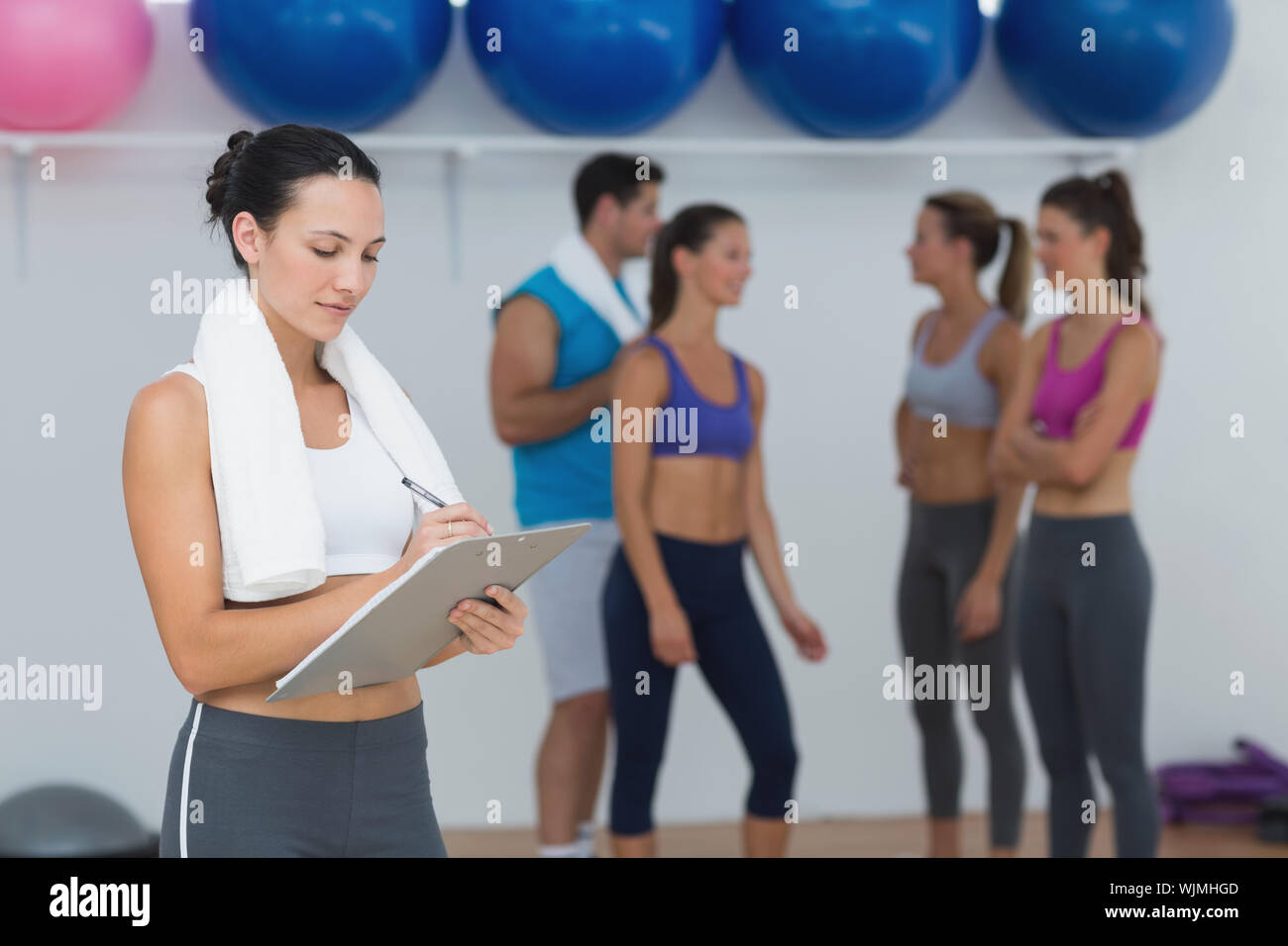 Female trainer writing on clipboard with fitness class in background at ...