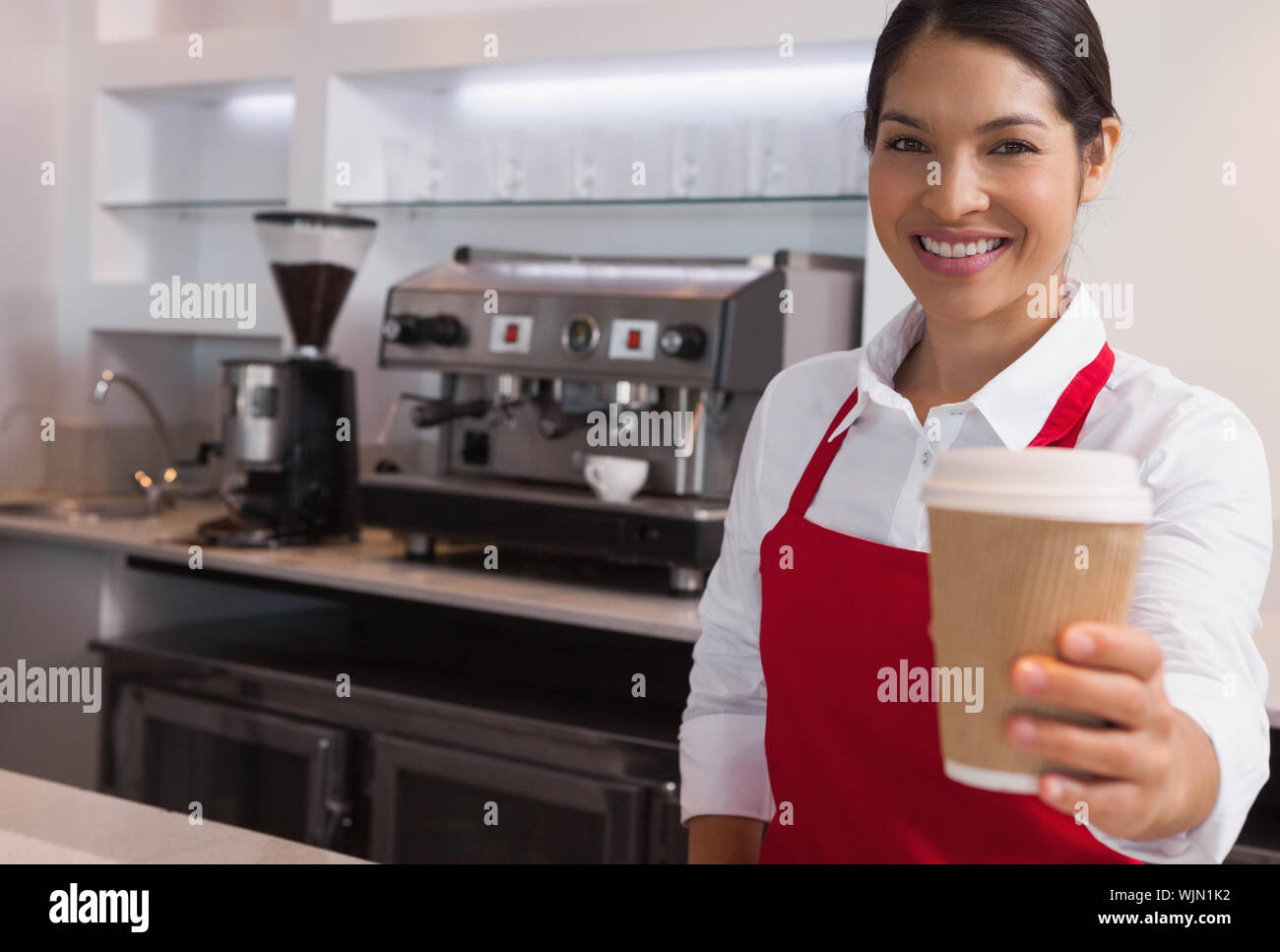 Happy young barista offering cup of coffee to go smiling at camera in a ...