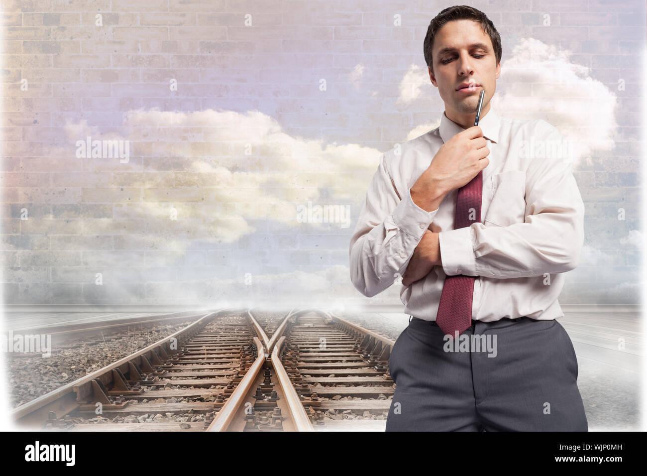 Thoughtful businessman holding pen to chin against train tracks leading ...