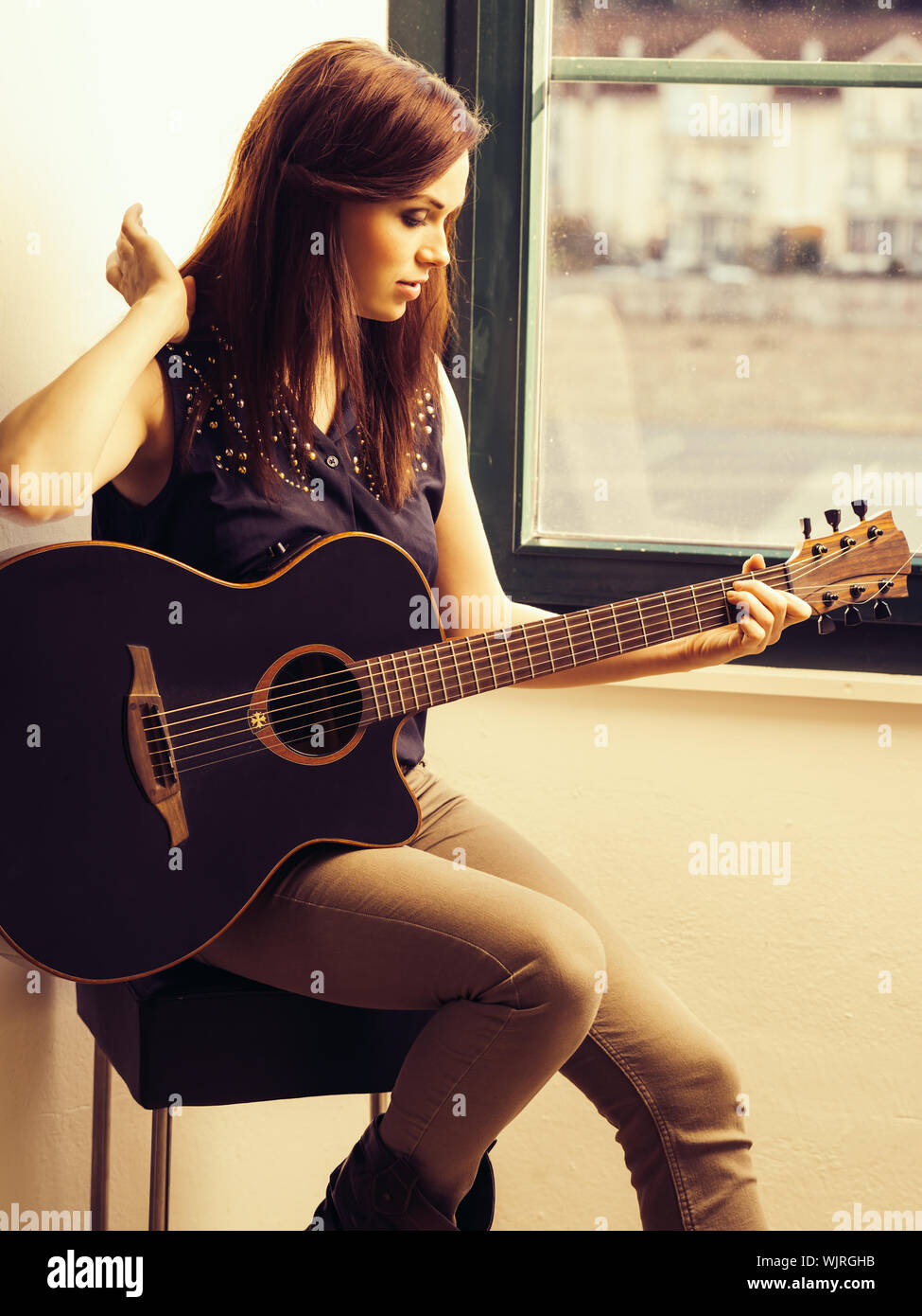 Photo of a woman playing an acoustic guitar while sitting by a window ...