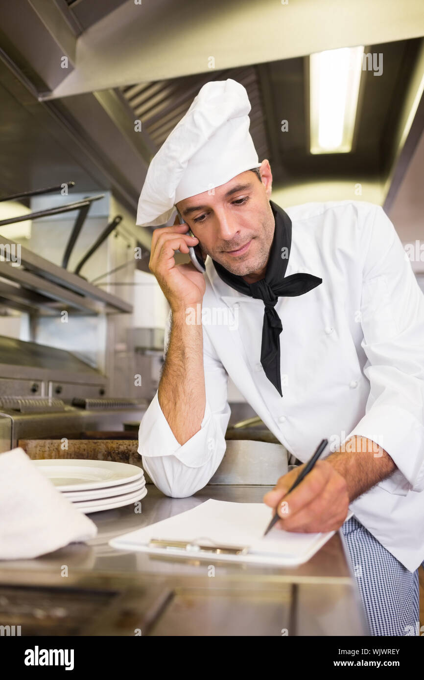 Concentrated male cook writing on clipboard while using cellphone in ...