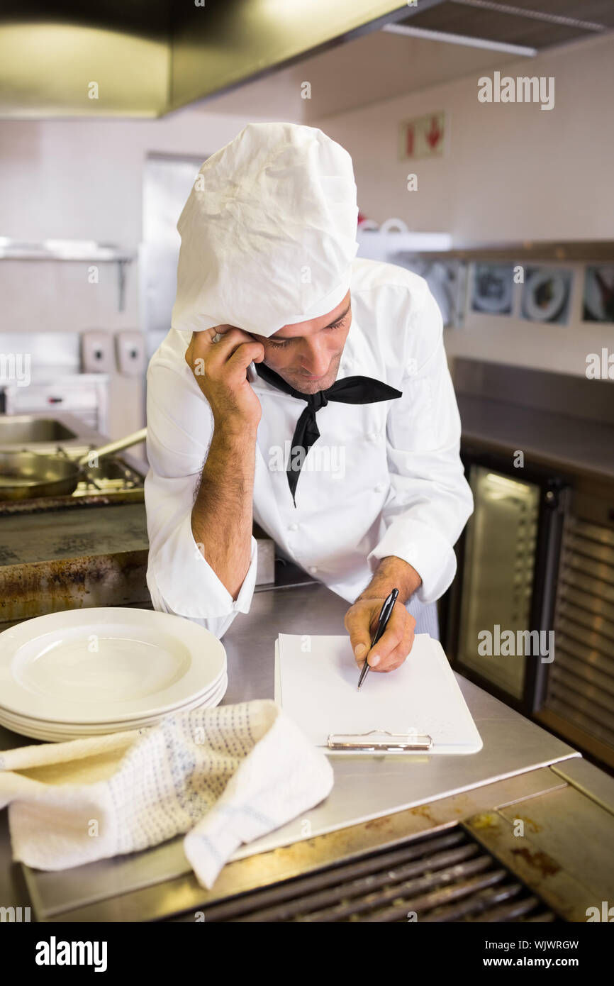 Concentrated male cook writing on clipboard while using cellphone in ...