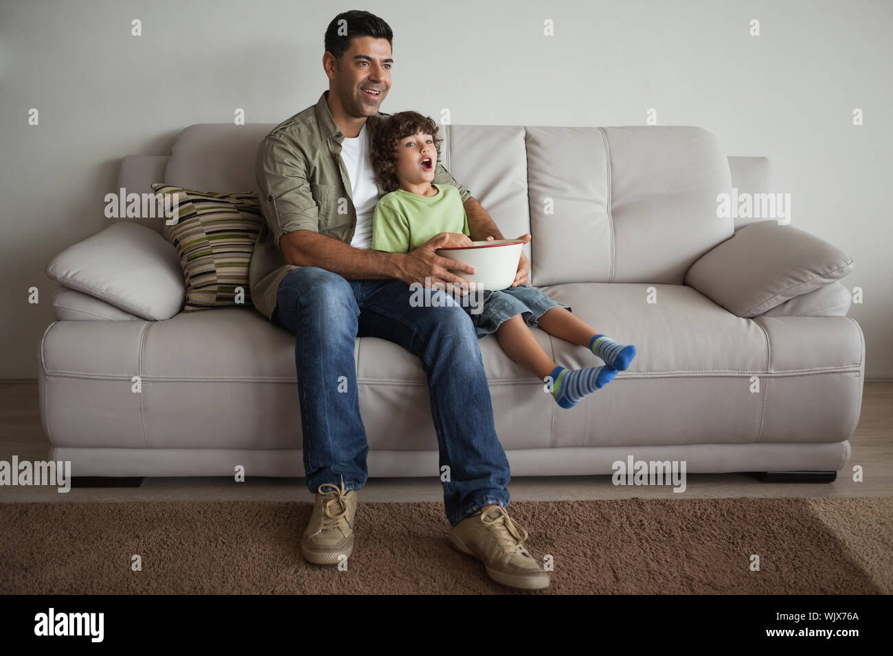 Full length of a happy father and son with popcorn bowl watching tv in ...