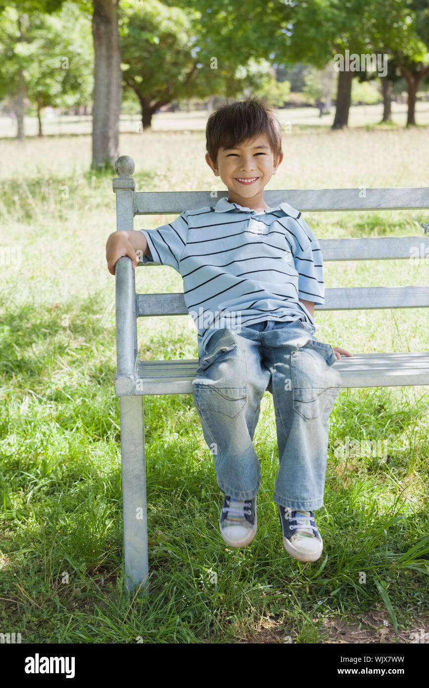 Full length portrait of a happy young boy sitting on bench at the park ...
