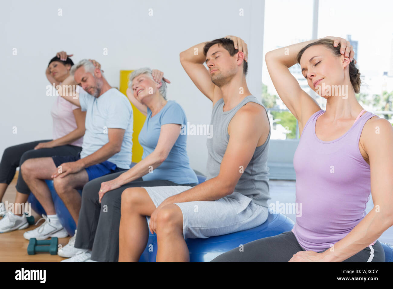 Fitness class sitting on exercise balls and stretching neck in a bright ...