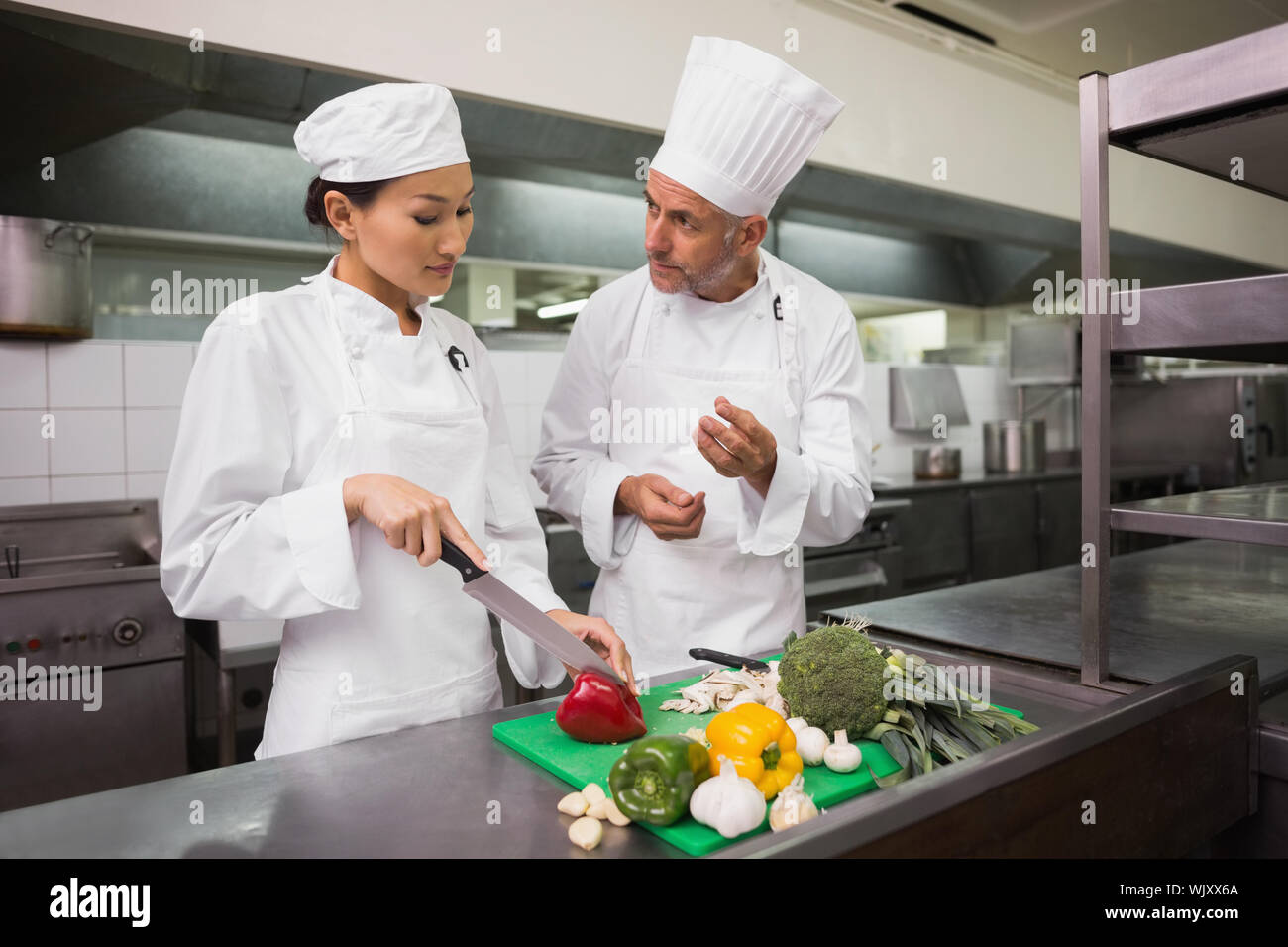 Chef teaching trainee how to slice vegetables in a commercial kitchen ...