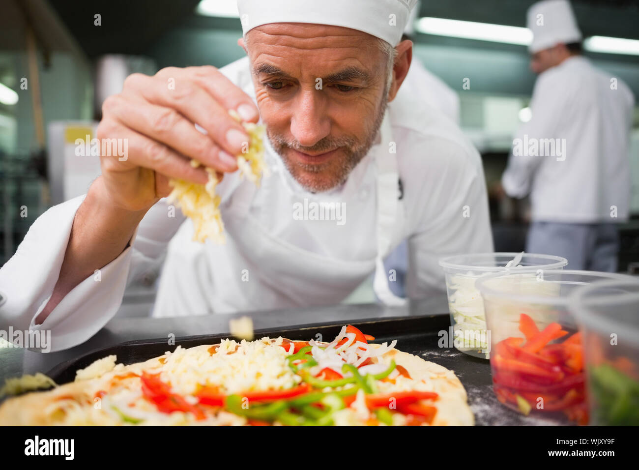 Happy chef sprinkling cheese on a pizza in a commercial kitchen Stock ...