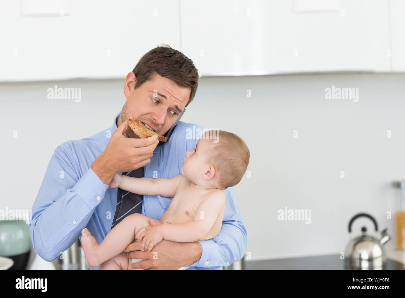 Busy father holding his baby son before work and eating toast at home ...