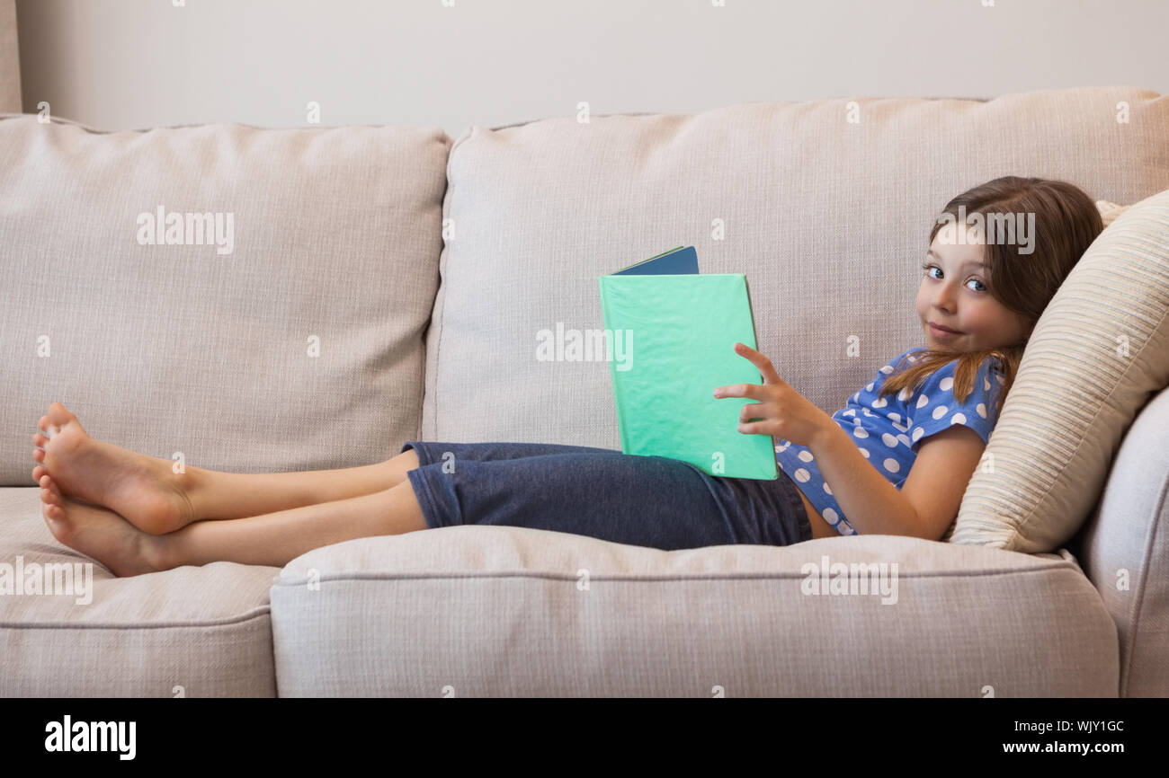 Full length portrait of a young girl reading a book on sofa at home ...