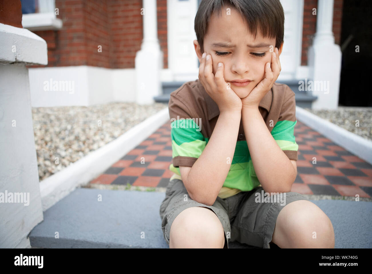 Sad little boy with hands on chin sitting on front steps of house Stock ...