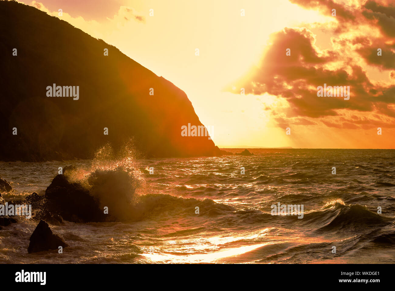 Golden light reflects on the water as waves break over rocky coastal ...