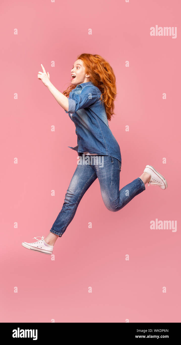 Redhead girl jumping and pointing finger aside on pink background Stock ...