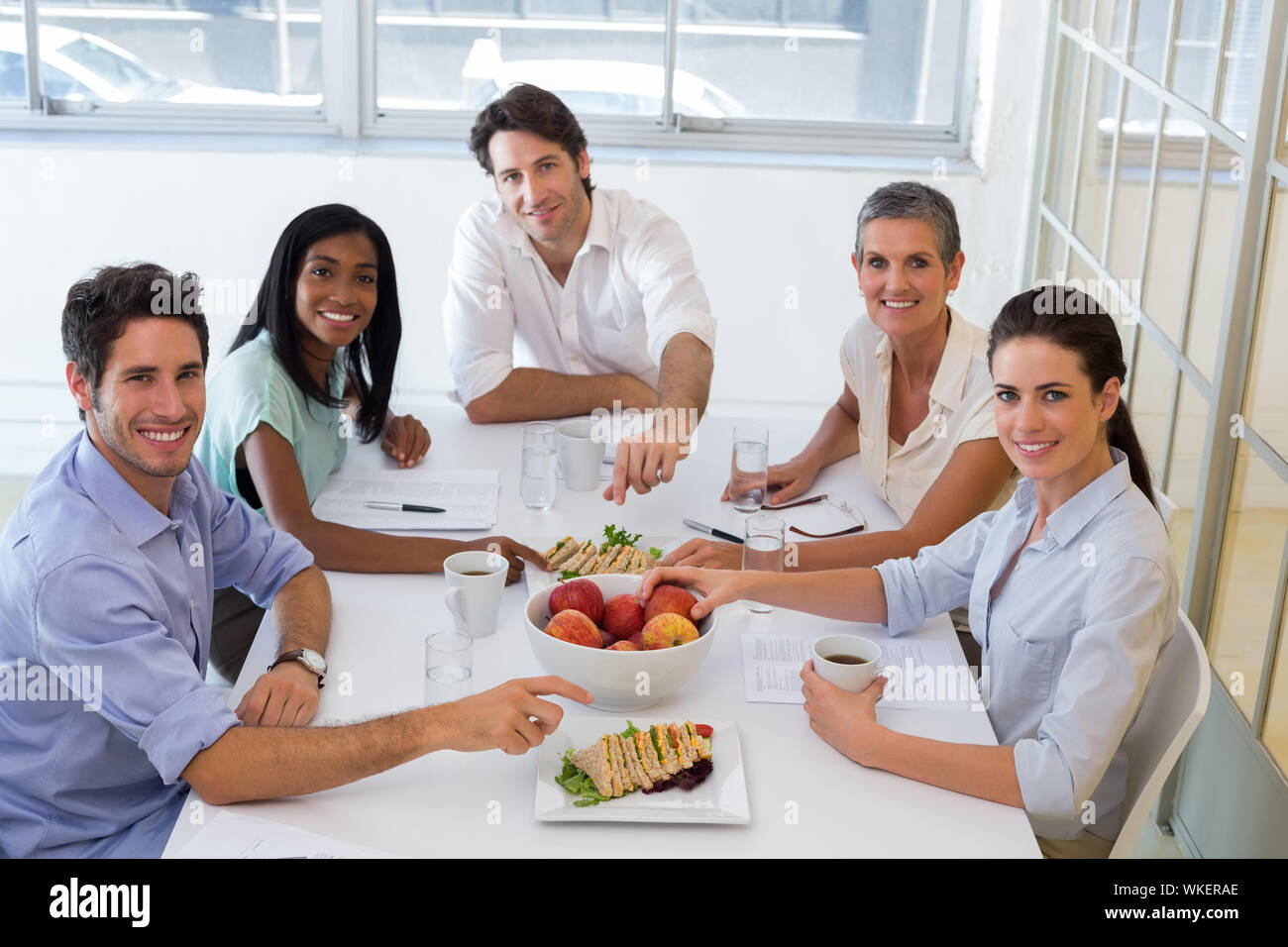 Business people smiling at camera eating sandwiches and fruit for lunch ...