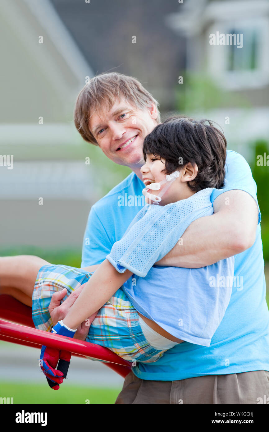 Father helping disabled seven year old son play on playground equipment ...