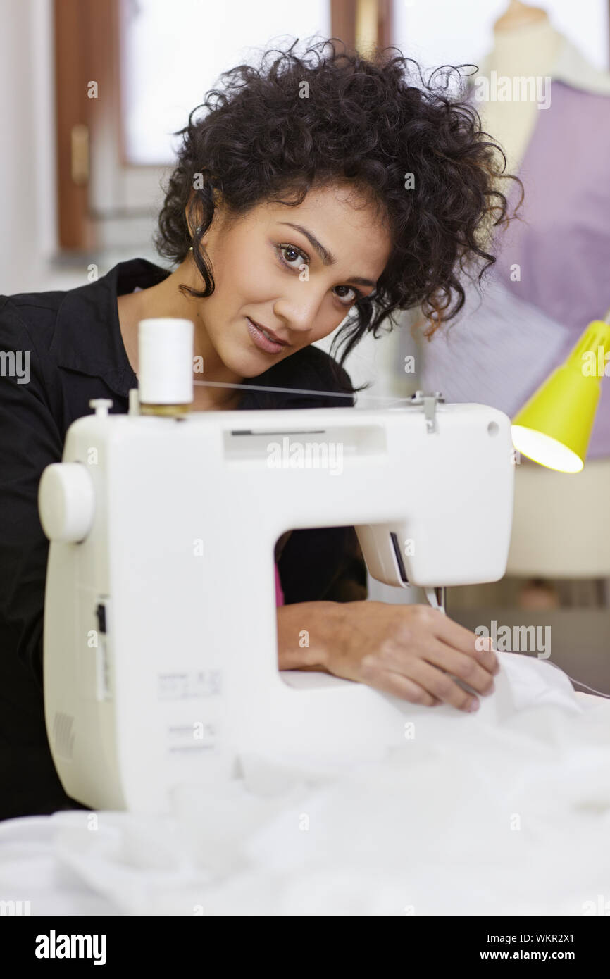 Young hispanic female dressmaker using sewing machine and smiling at ...