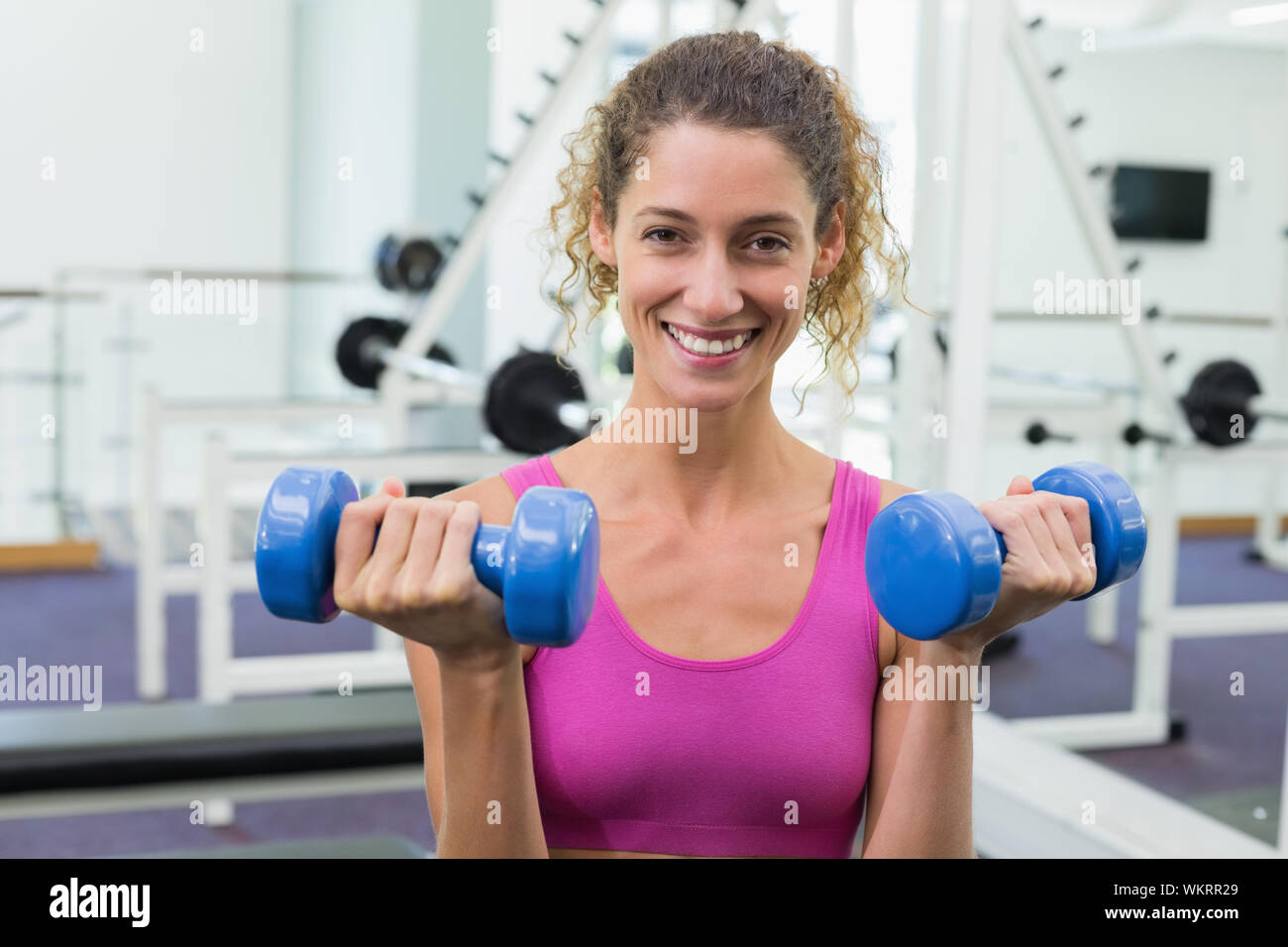 Pretty fit woman lifting blue dumbbells smiling at camera at the gym ...