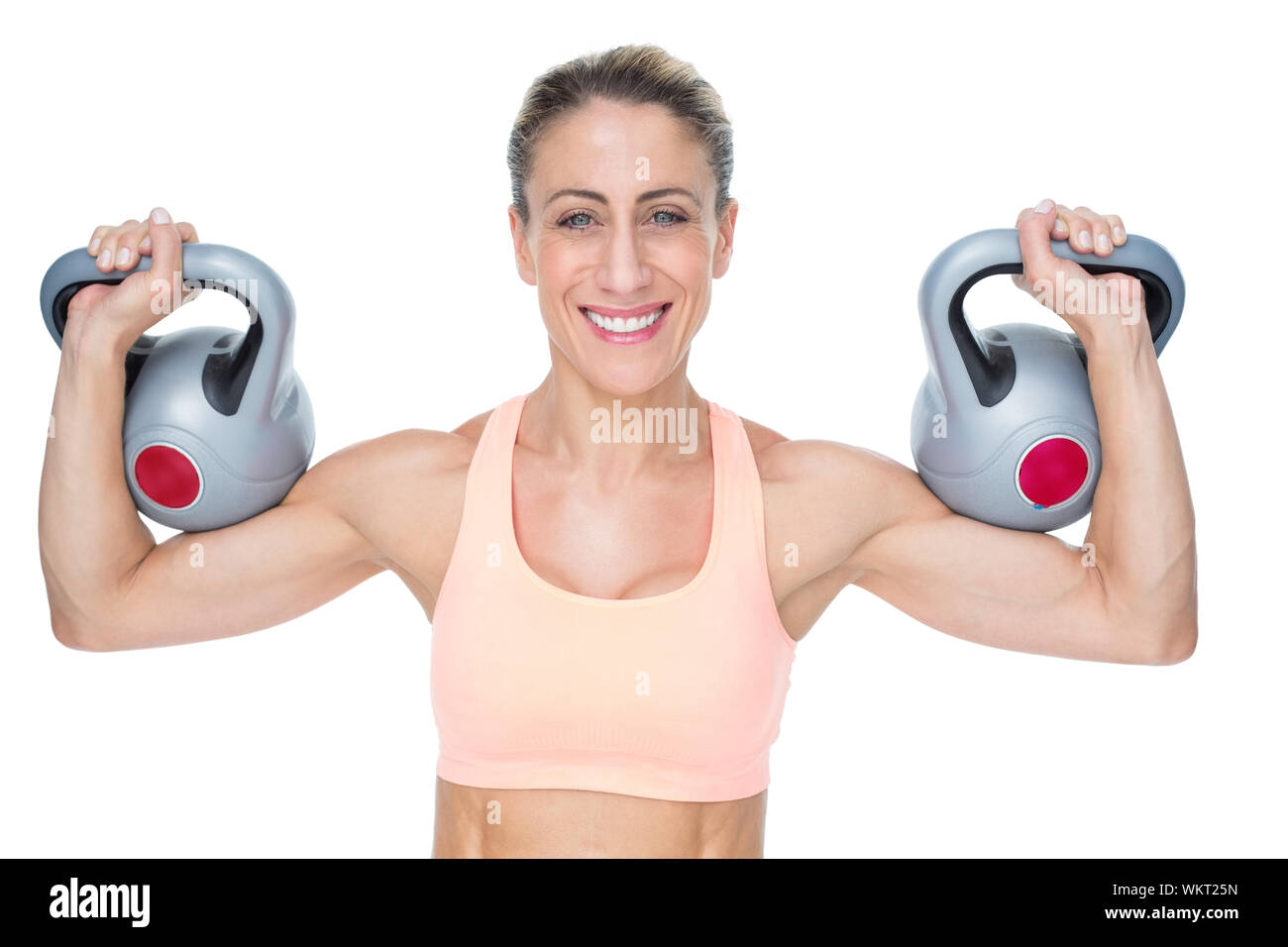 Happy female crossfitter lifting kettlebells looking at camera on white ...