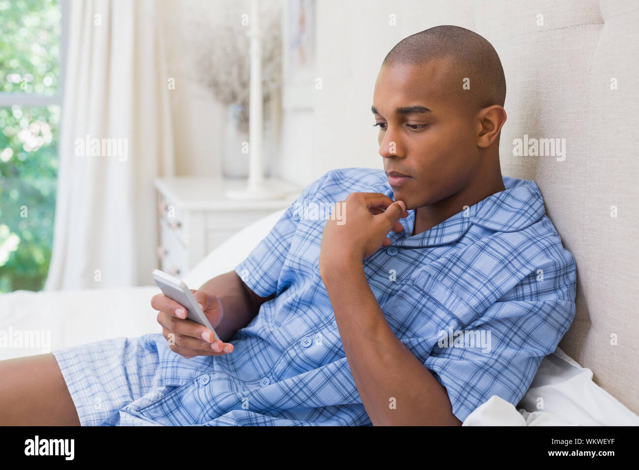 Thinking man sitting on bed and texting on phone at home in the bedroom ...
