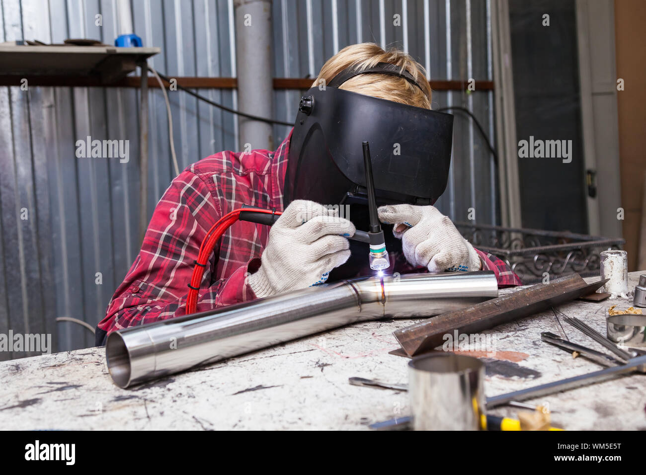 Young guy welder in a checkered red shirt welds a stainless steel pipe ...