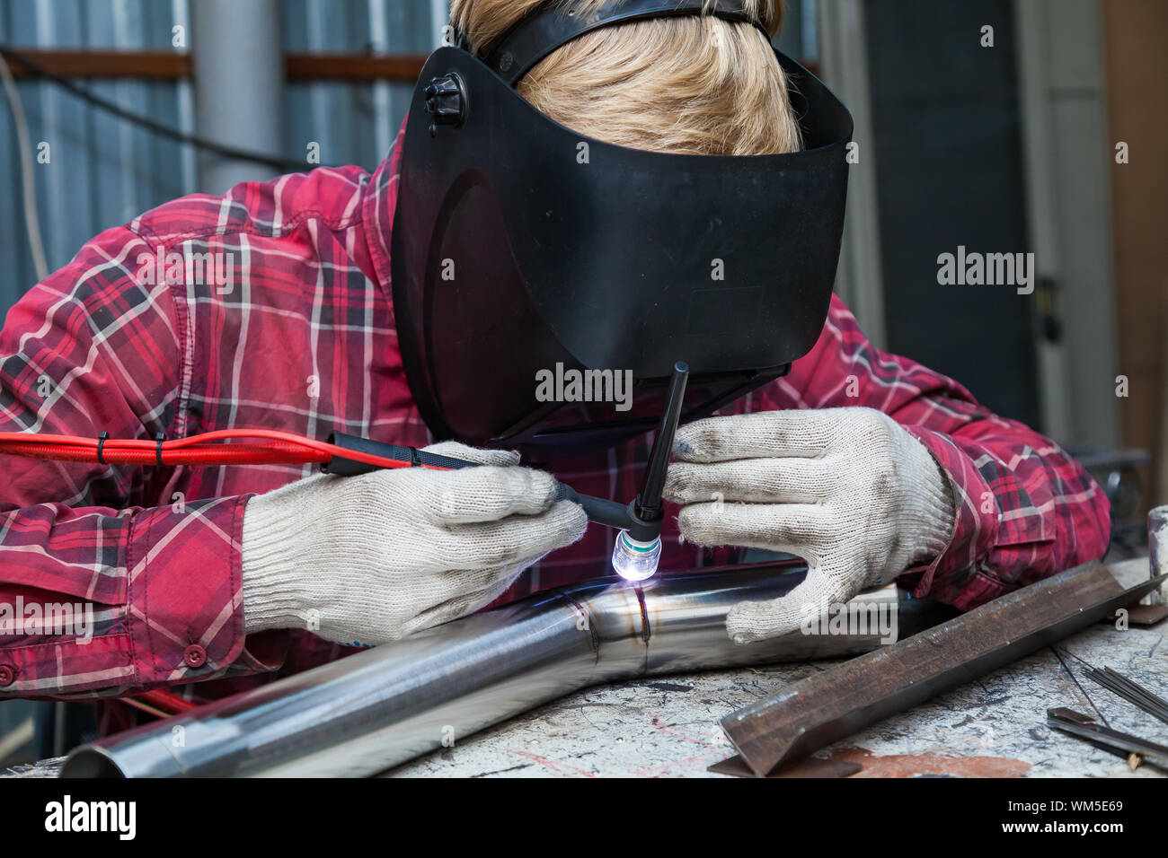 Young guy welder in a checkered red shirt welds a stainless steel pipe ...