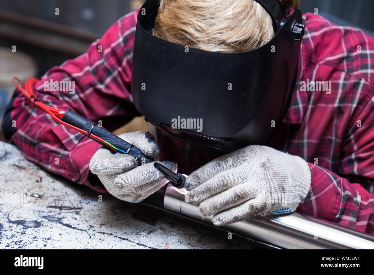Young guy welder in a checkered red shirt welds a stainless steel pipe ...