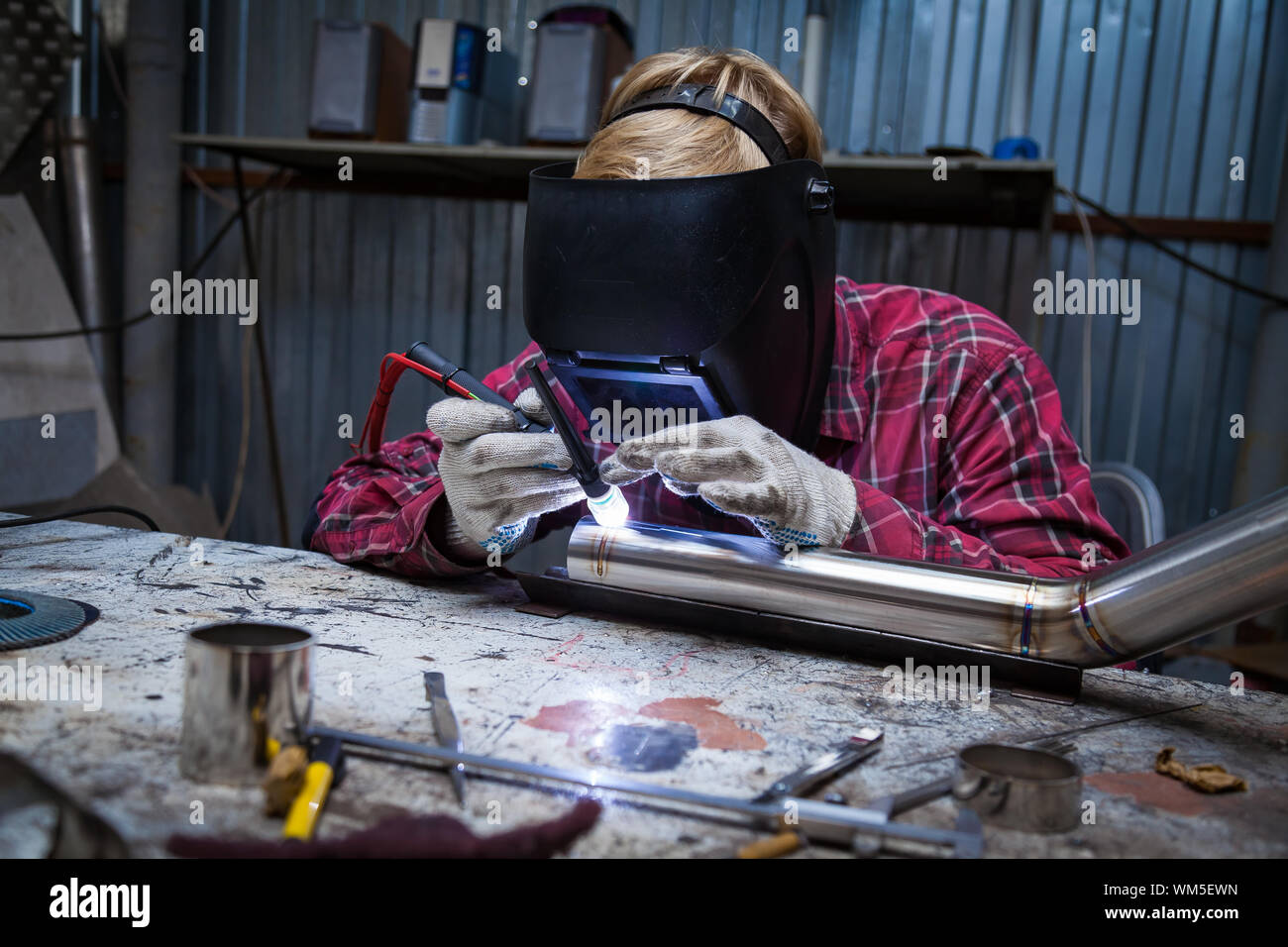 Young guy welder in a checkered red shirt welds a stainless steel pipe ...