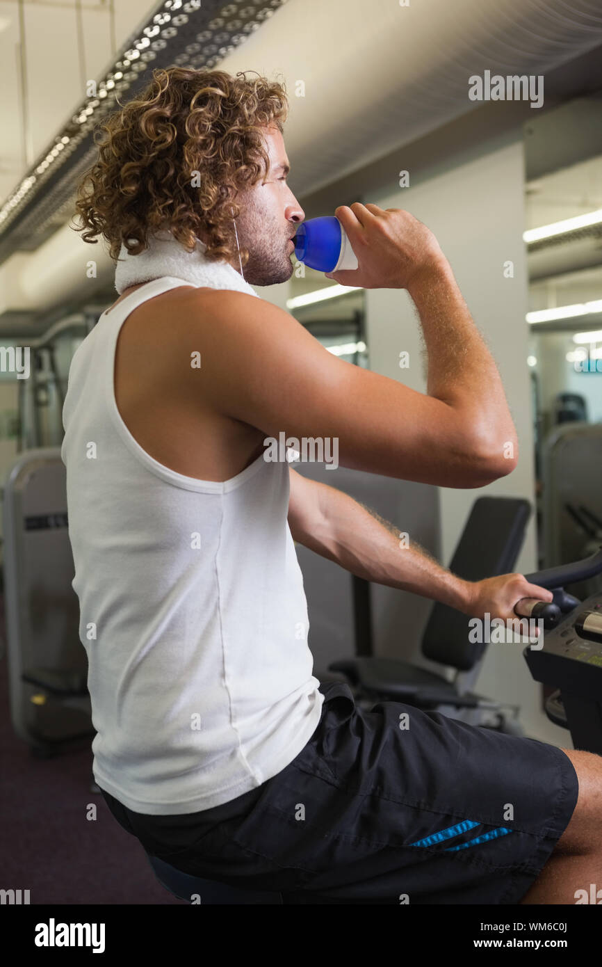 Side view of a young man drinking water on exercise bike at the gym ...