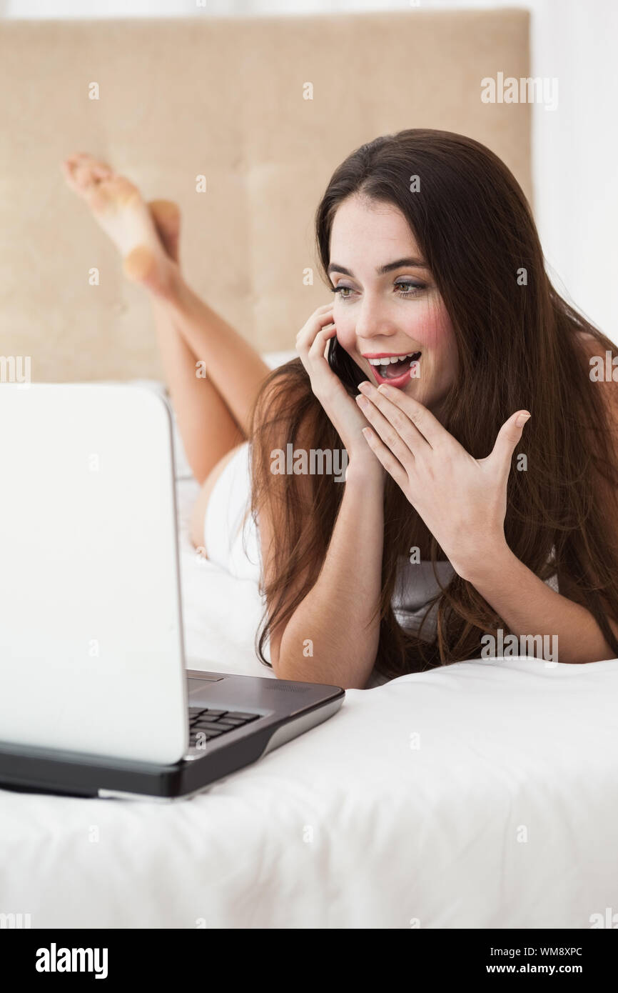 Pretty brunette on the phone in bed using laptop at home in the bedroom ...