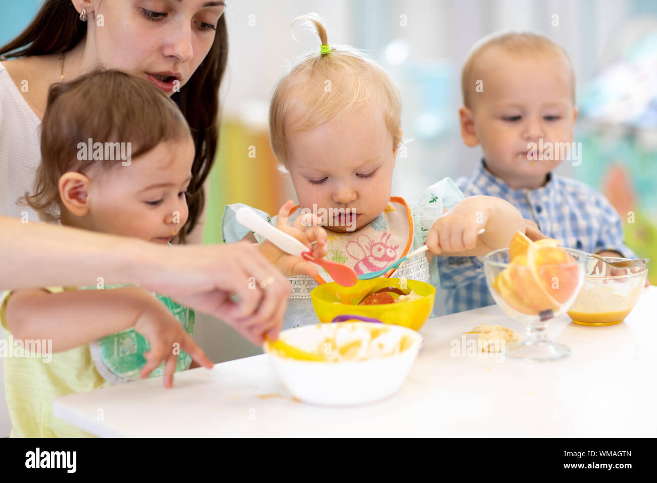 Group of nursery babies eating healthy food with kindergartener help ...