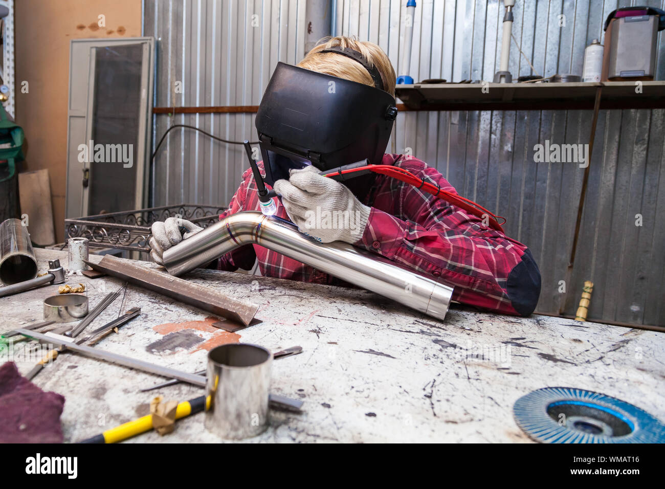 Young guy welder in a checkered red shirt welds a stainless steel pipe ...