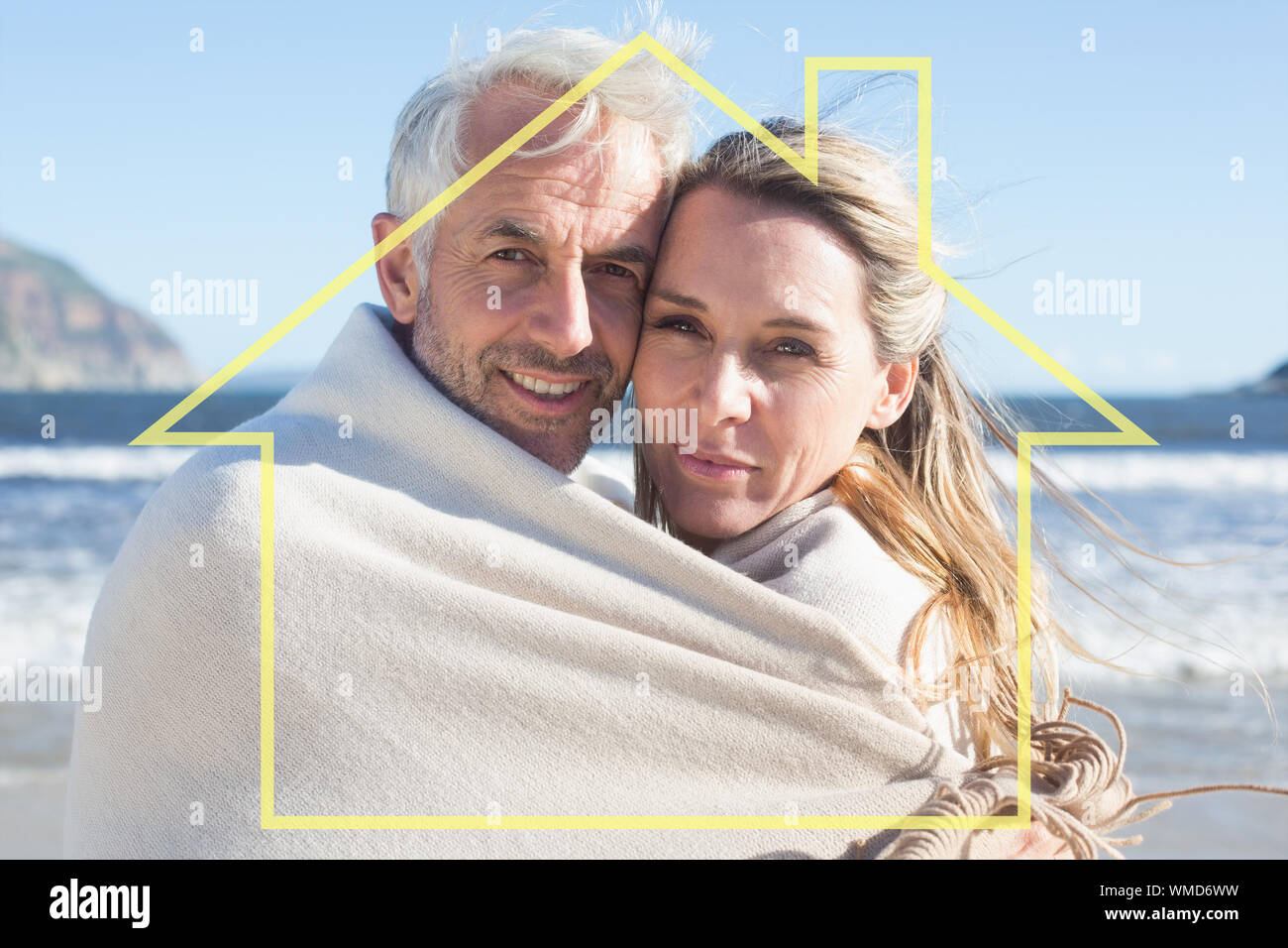 Smiling couple wrapped up in blanket on the beach against house outline ...