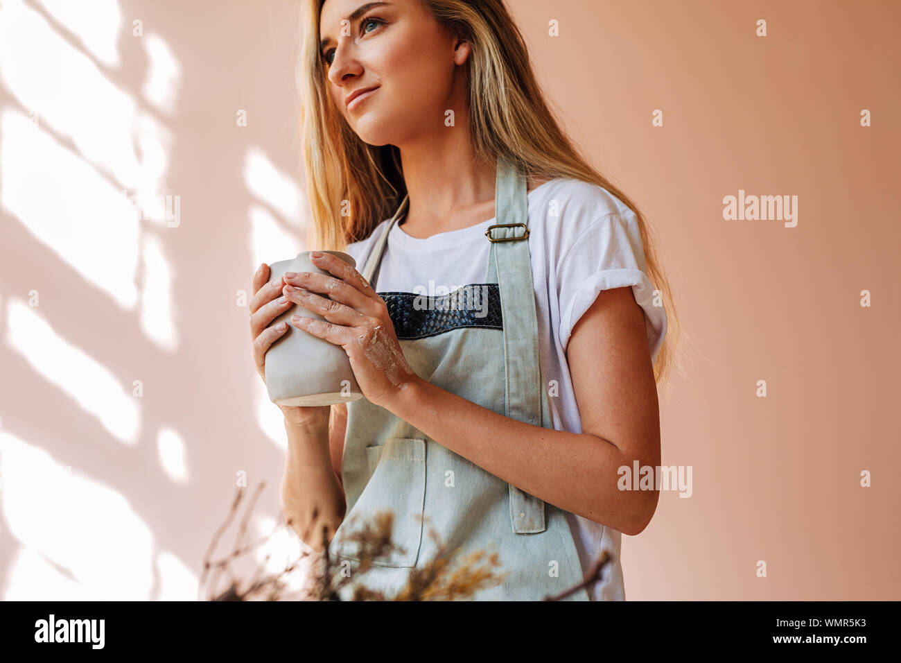 Side view of female ceramic artist in apron carefully holding clay mug ...