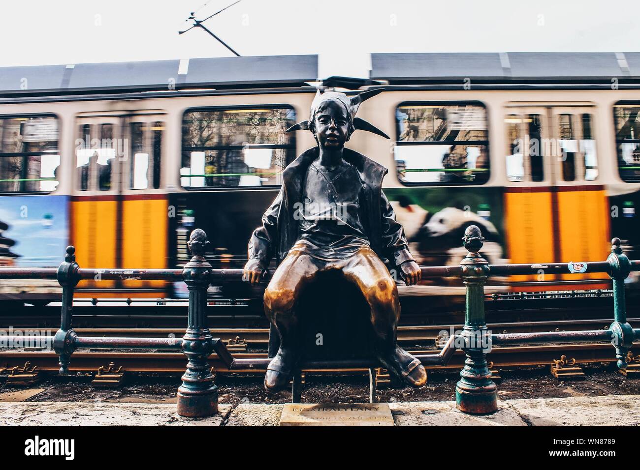 Close shot of a kid's statue sitting on a metal fence with a train in ...