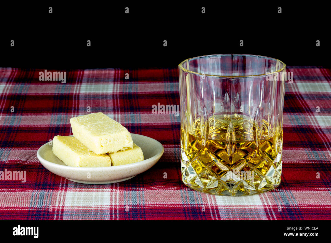 Whisky and shortbread on tartan table cloth against black background ...