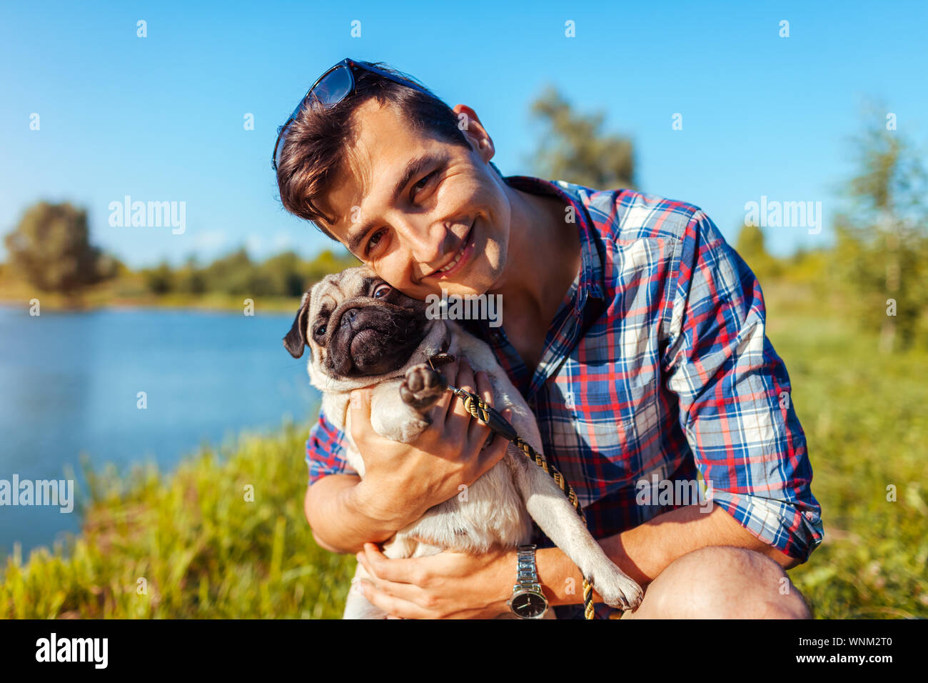 Man hugging pug dog in park. Happy man walking with pet by summer lake ...