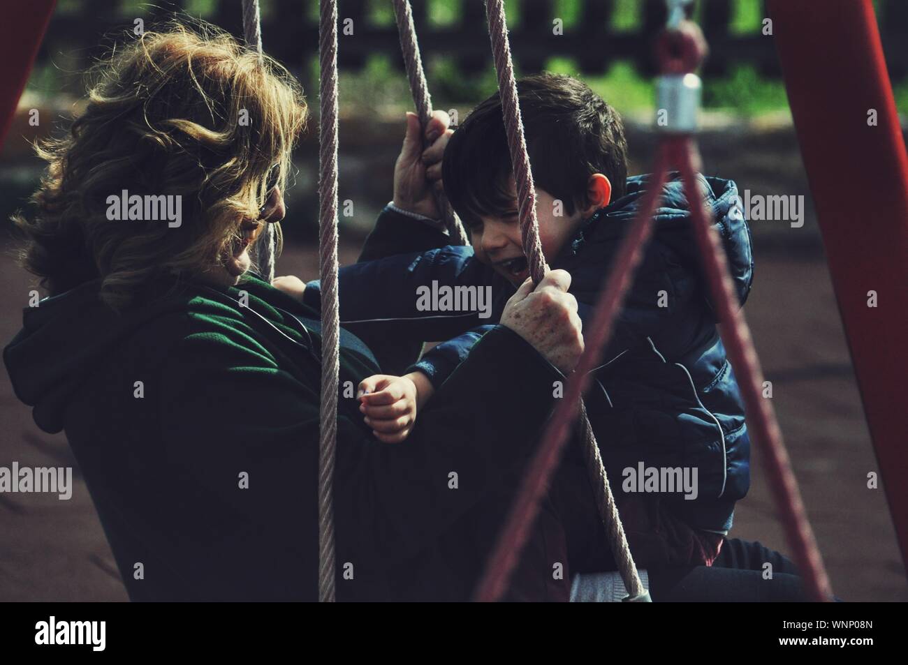 Side View Of Grandmother With Grandson Sitting On Swing At Playground