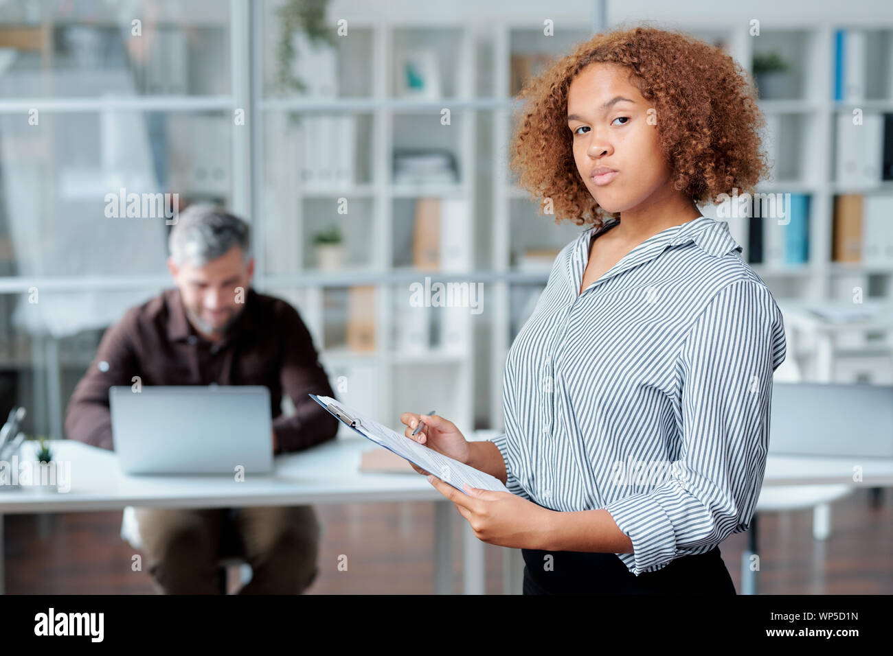 Serious young intercultural secretary with pen and clipboard reading ...
