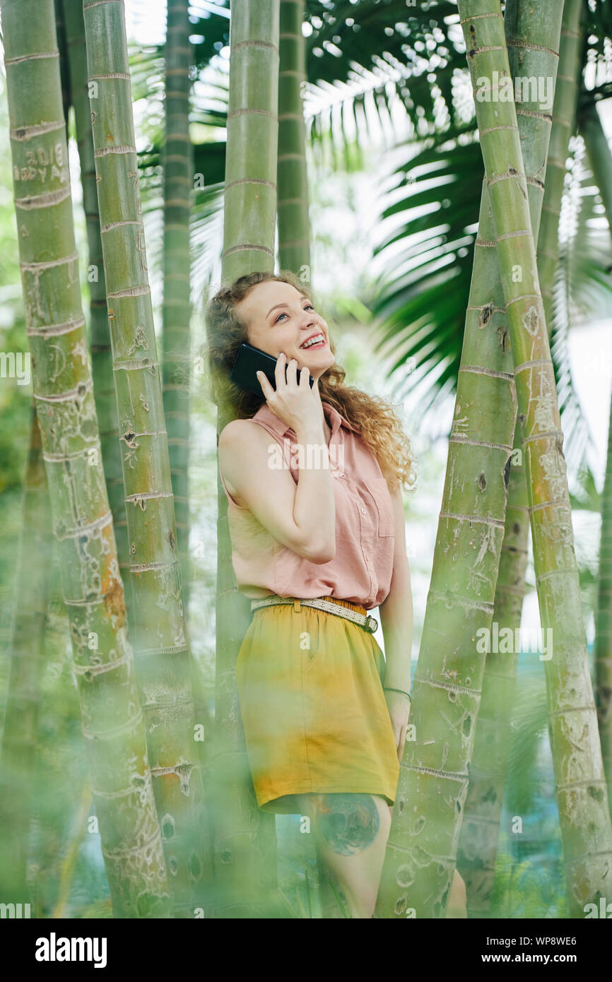 Happy young woman leaning on bamboo tree and talking on phone with ...