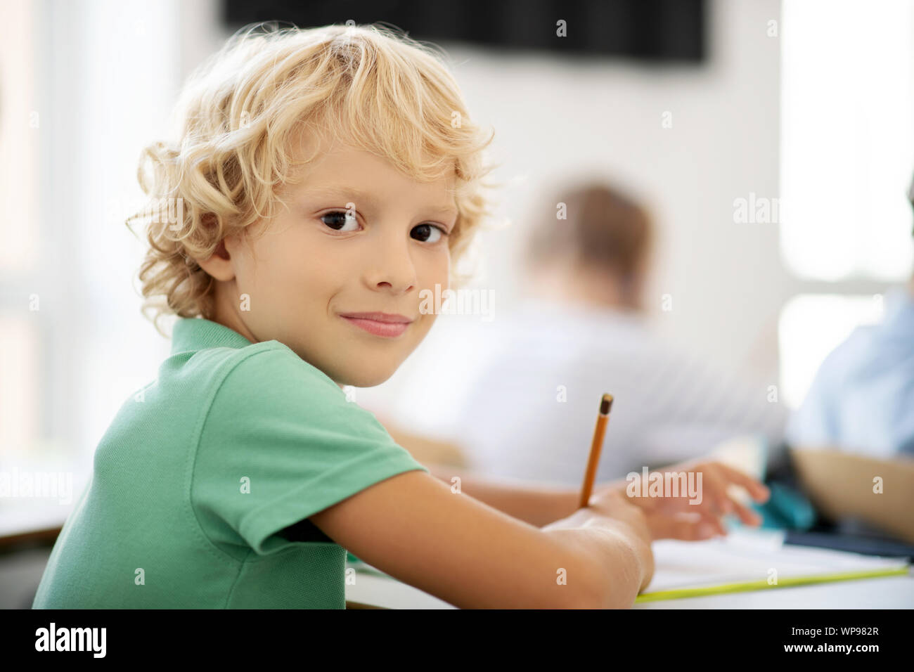 Handsome curly boy holding pencil while sitting at the desk Stock Photo ...