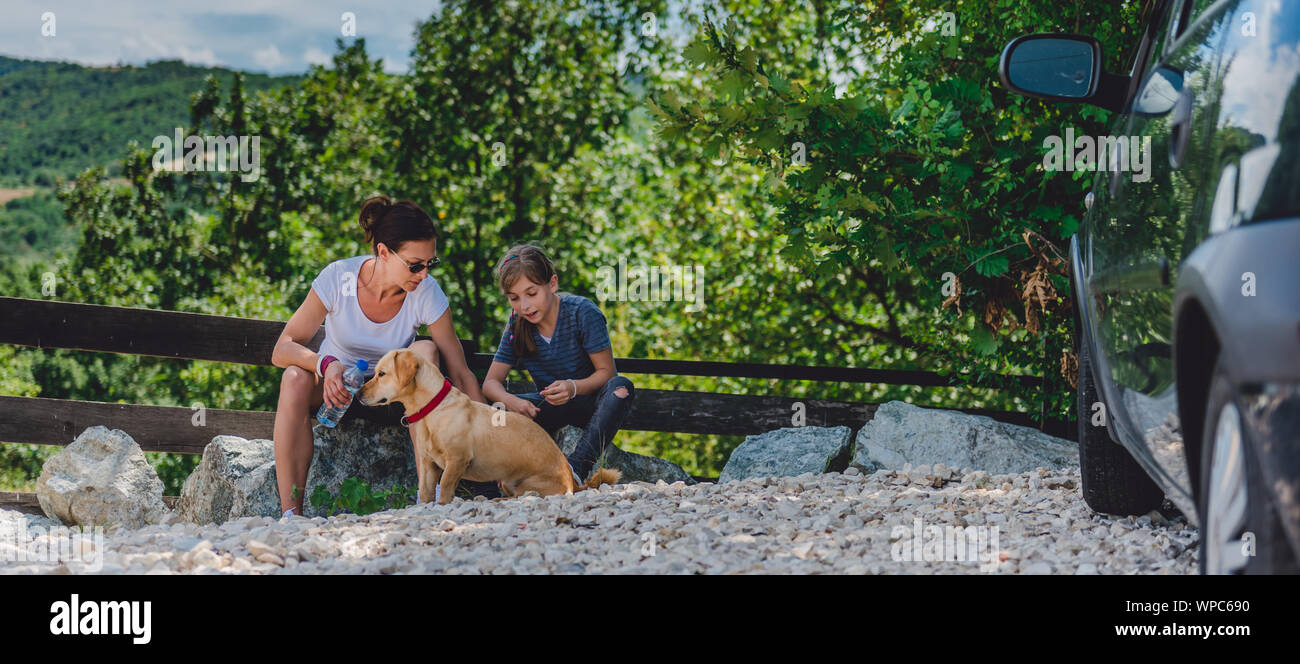 Mother and daughter on a road trip with dog taking a break and sitting ...