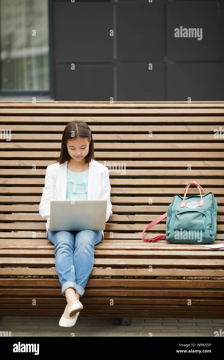 Asian schoolgirl with backpack sitting on the bench and communicating ...