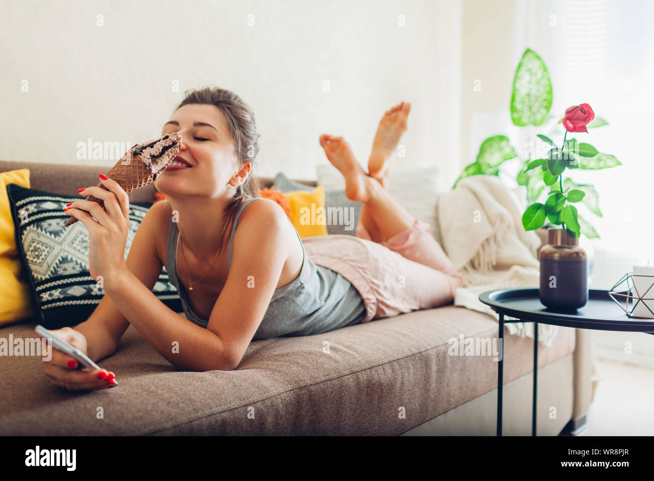 Happy young woman eating chocolate ice-cream in cone lying on couch at ...