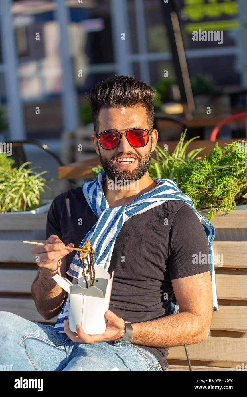 Young male hipster with a mustache and beard is eating chinese noodles ...
