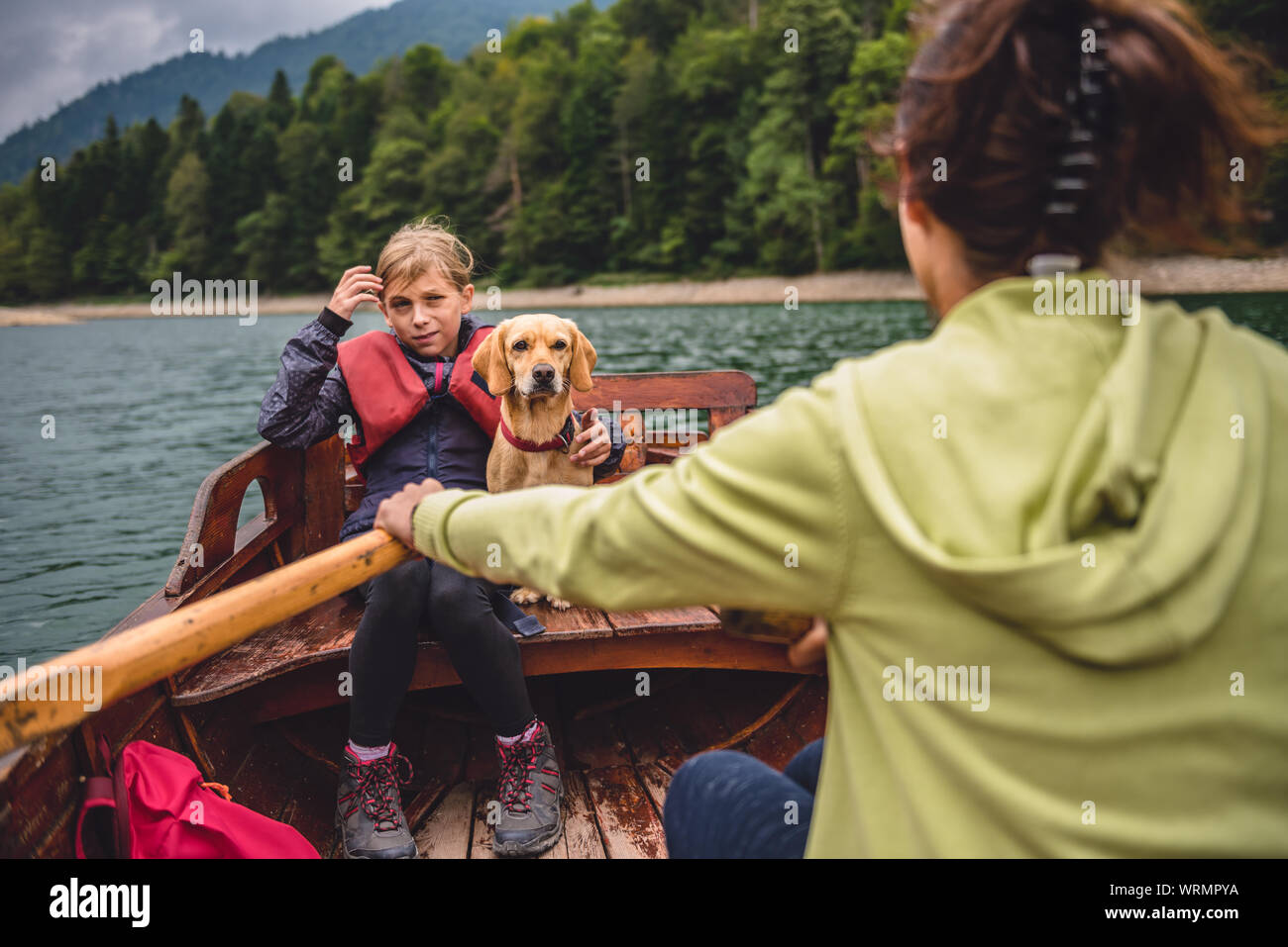 Mother and daughter with a small yellow dog rowing a boat on a mountain ...