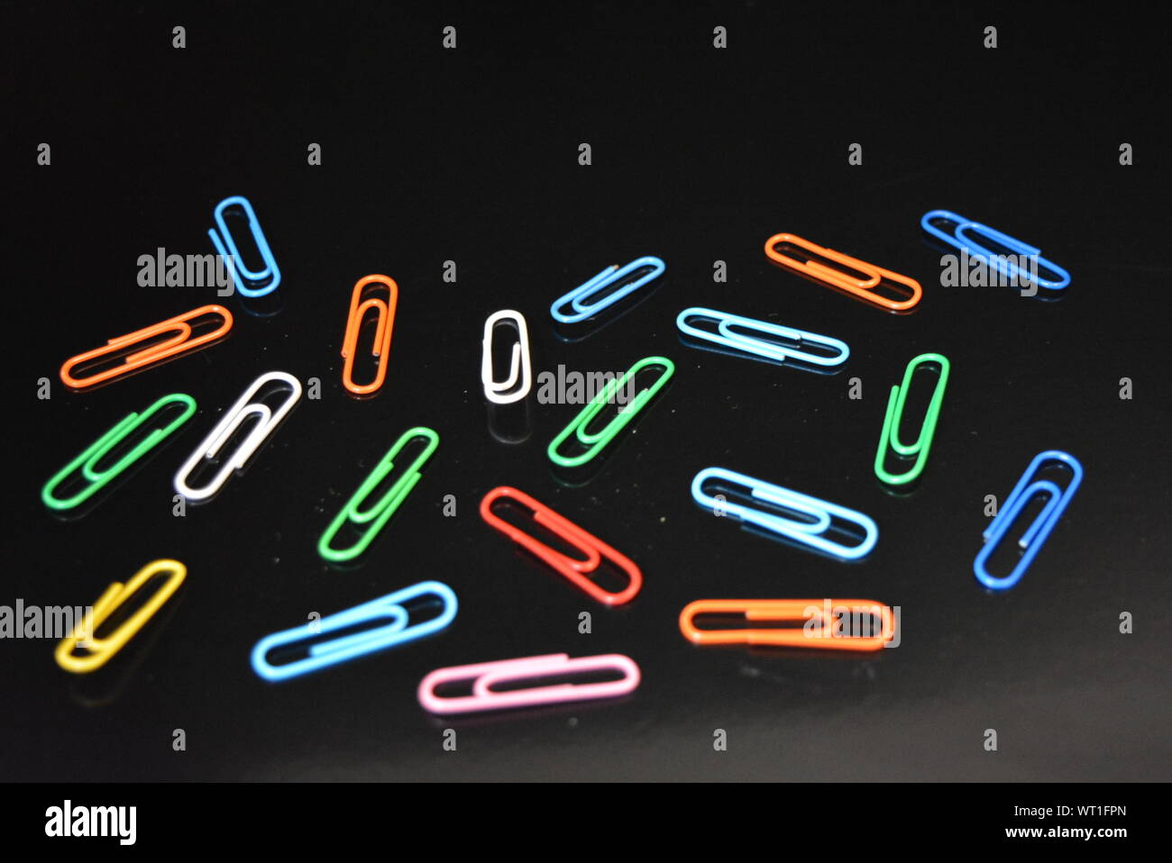 Many bright glossy colorful paper clips lie on a black glossy surface ...