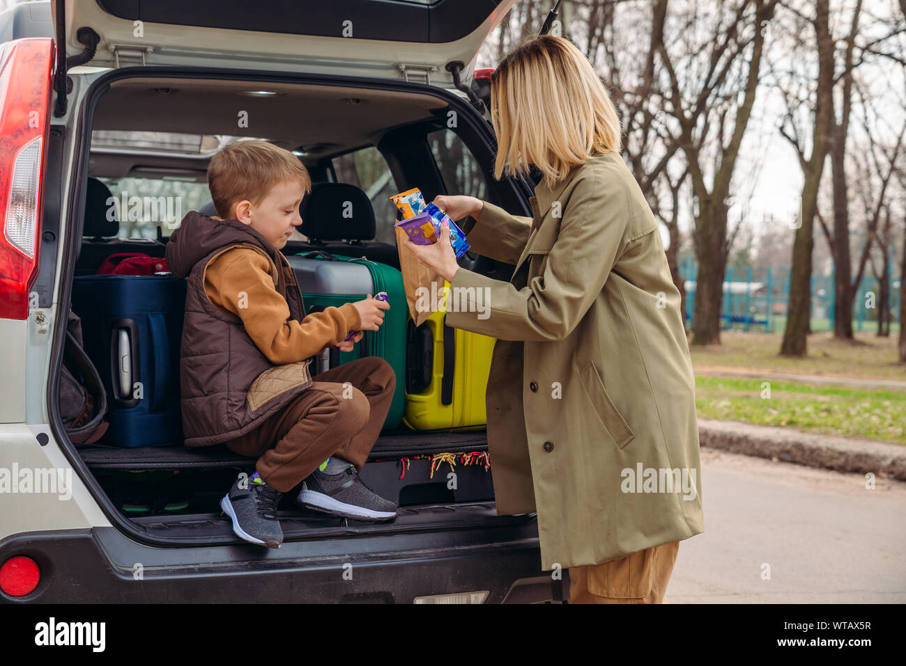 little kid sitting with mother in car trunk full of luggage eating ...