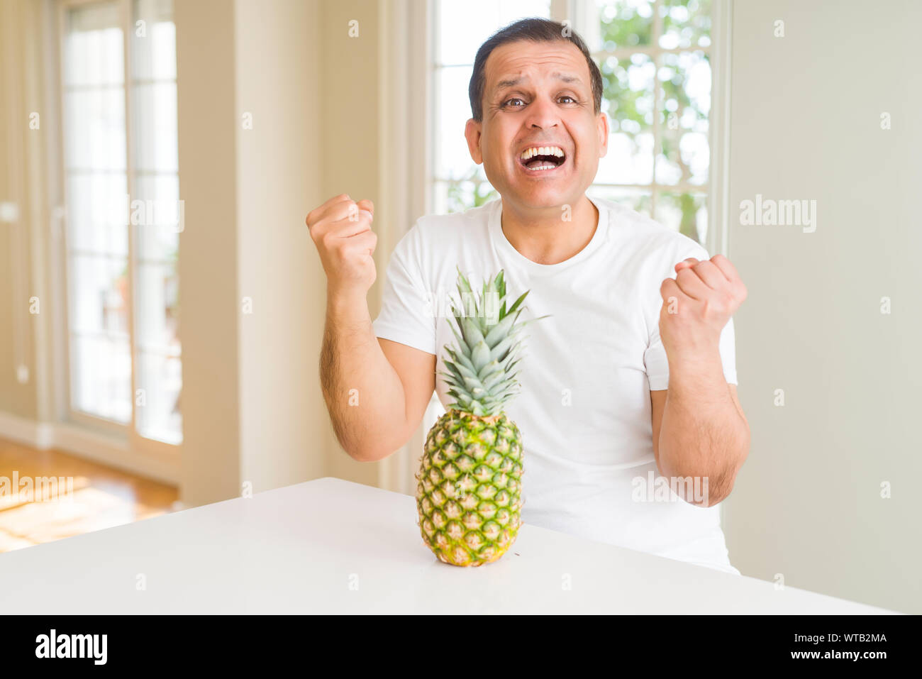 Middle age man eating fresh tropical pineapple at home screaming proud ...