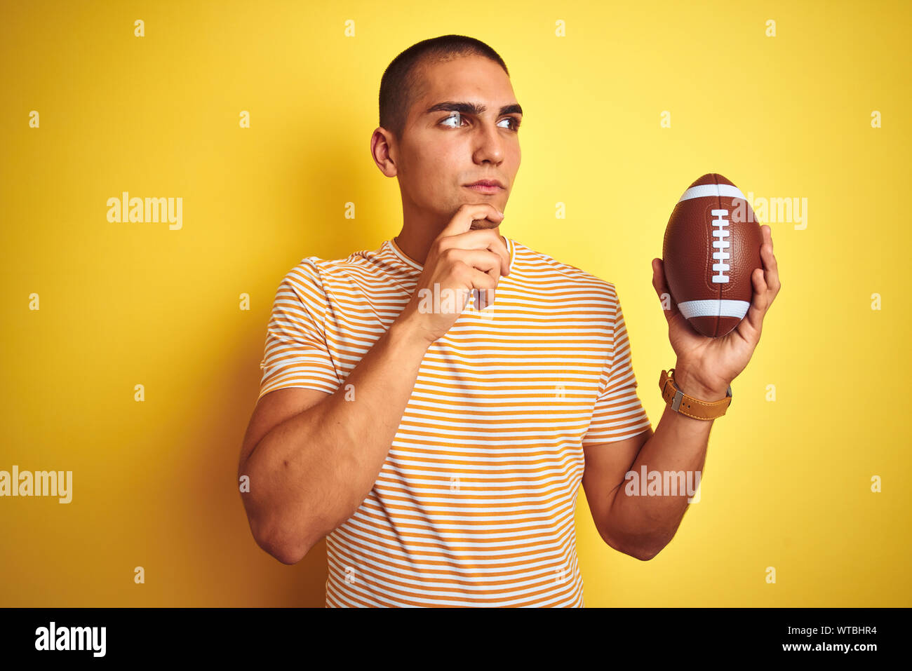 Young rugby player man holding a football ball over yellow isolated ...